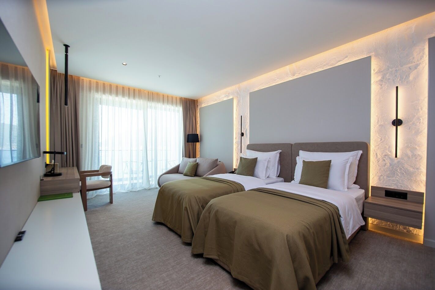 Quarto de hotel com duas camas, uma escrivaninha, uma cadeira, televisão e uma janela grande com cortinas.