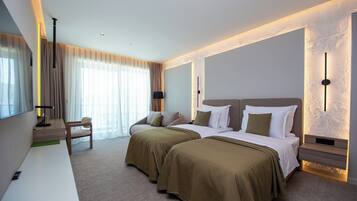Quarto de hotel com duas camas, uma escrivaninha, uma cadeira, televisão e uma janela grande com cortinas.