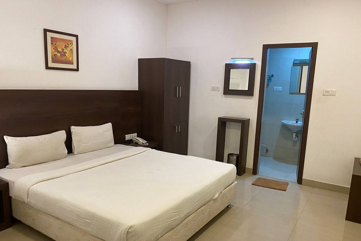 Quarto de hotel com uma cama grande, um guarda-roupa, uma mesa de cabeceira com telefone, um quadro na parede e um banheiro visível por uma porta aberta.
