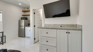 Uma cozinha moderna com armários brancos, bancada de granito, TV de tela plana montada acima dos armários e uma geladeira ao fundo.