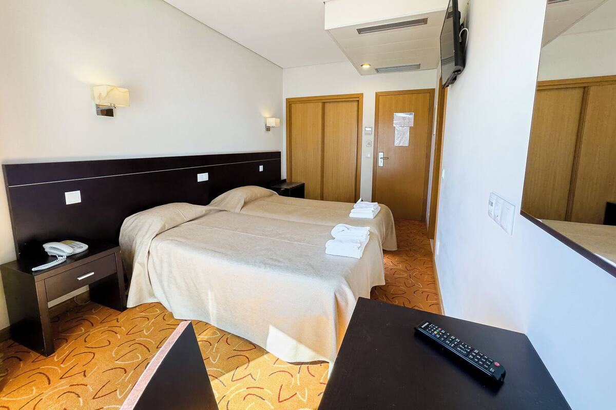 Quarto de hotel com duas camas, uma escrivaninha, uma cadeira, uma TV e telefone na mesinha de cabeceira.