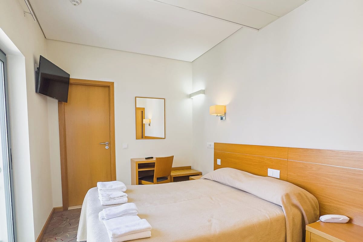 Quarto de hotel com cama, mesa de cabeceira, televisão e escrivaninha com cadeira.