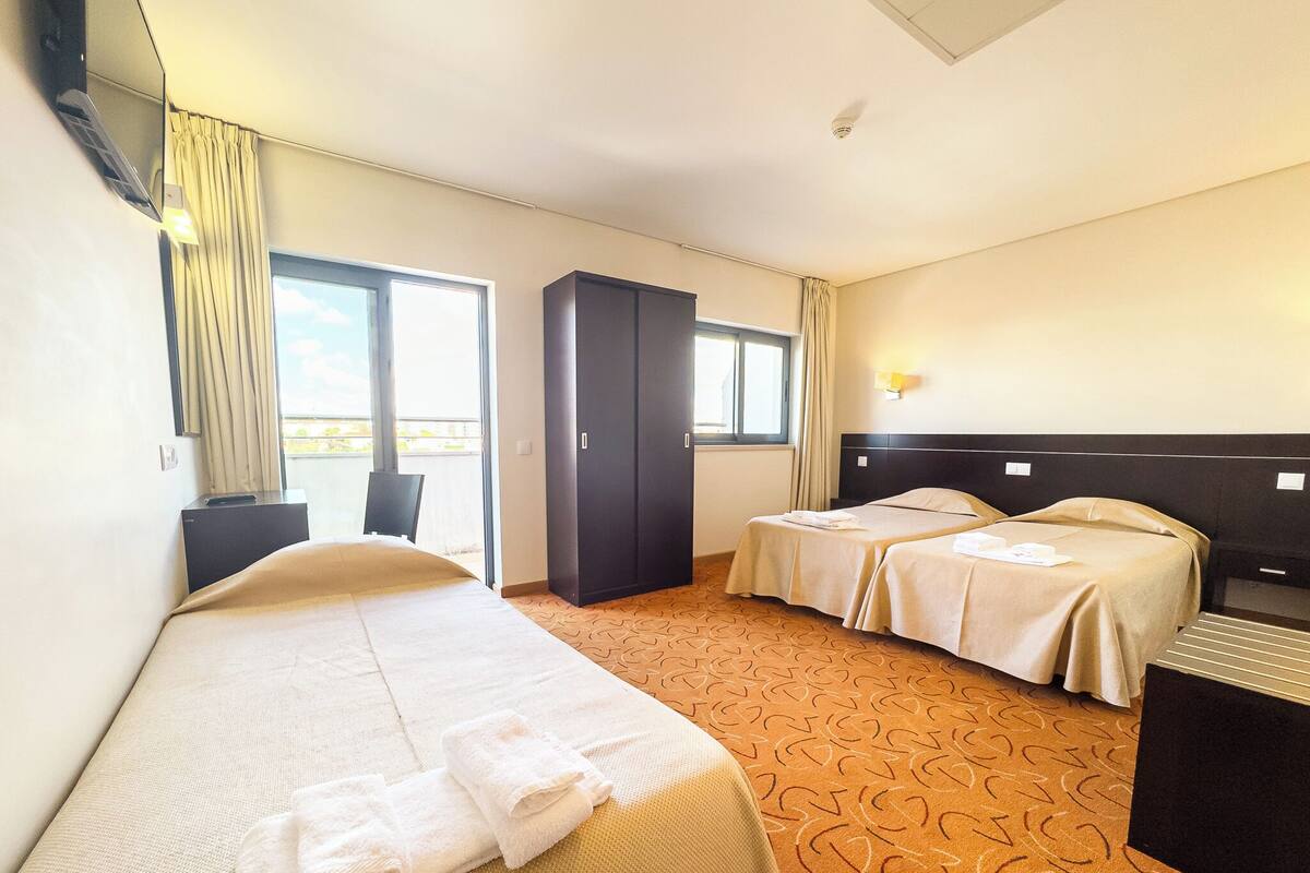 Quarto de hotel com duas camas, um guarda-roupa, uma escrivaninha e uma janela com cortinas.