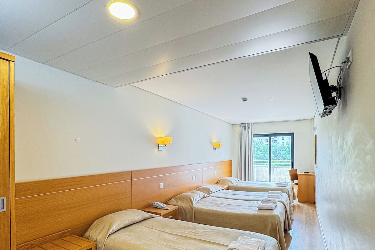 Quarto de hotel com três camas, cabeceiras de madeira, um banco de madeira, uma escrivaninha e uma televisão fixada na parede.