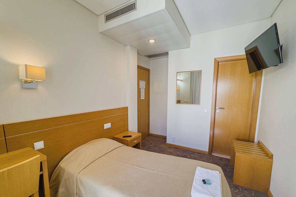 Quarto de hotel com uma cama, uma escrivaninha, uma cadeira, uma TV fixada na parede e um guarda-roupa de madeira.