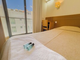 Quarto de hotel com uma cama, janela, uma cadeira e duas garrafas sobre a cama.