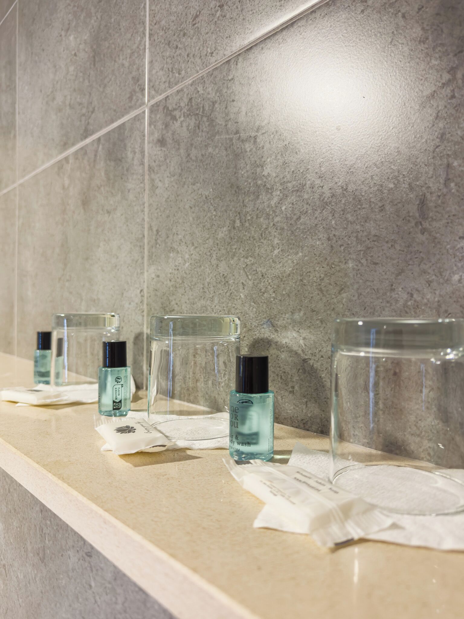 Um banheiro com parede revestida de azulejos, bancada e várias garrafas de vidro transparentes com sabonete líquido.
