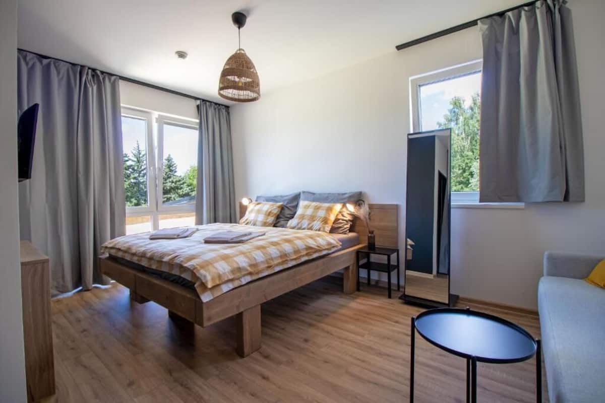 Quarto com cama, mesinha, cadeira, televisão e janela com cortinas.