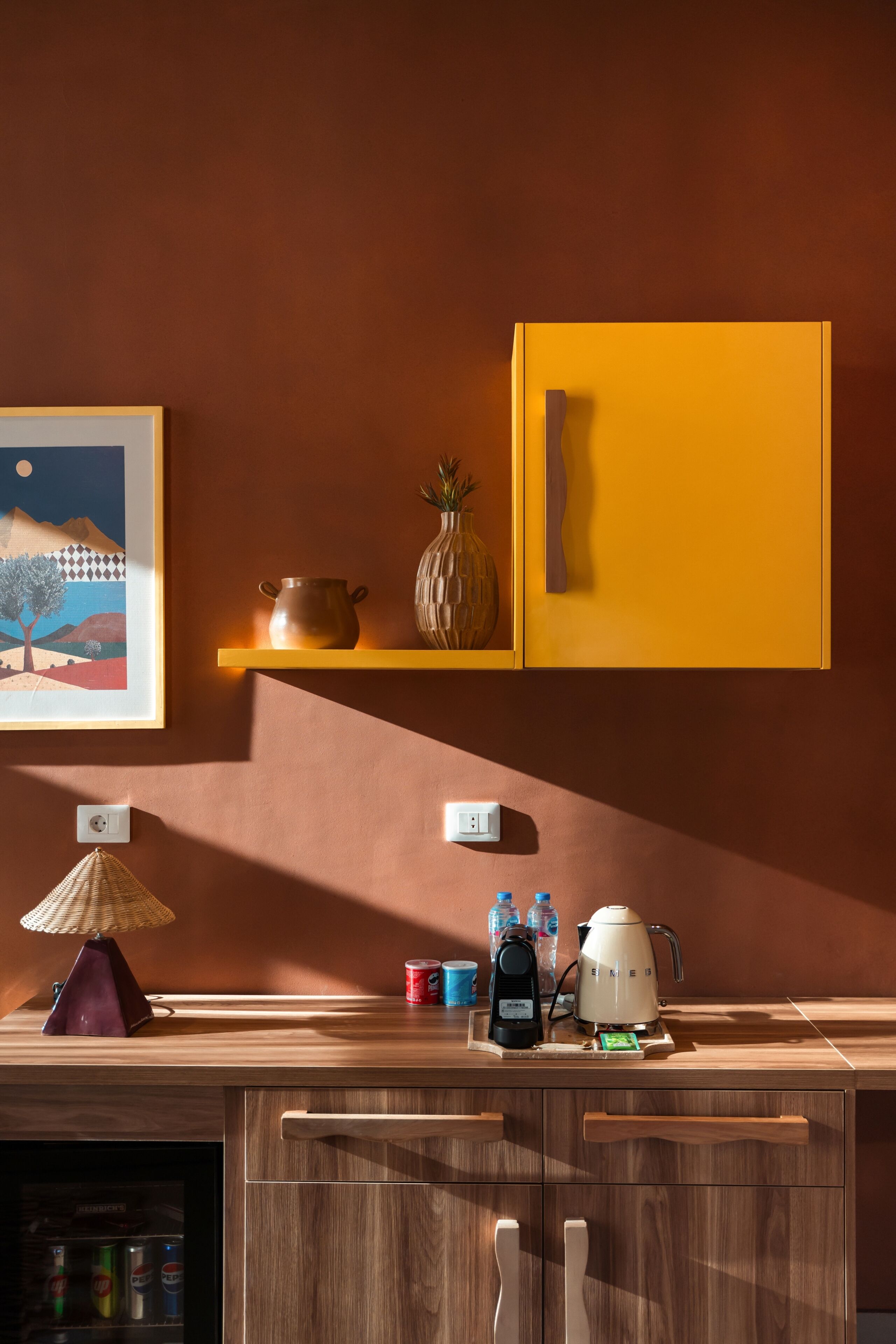 Uma cozinha com bancada de madeira, um armário amarelo, um quadro pendurado, uma prateleira com uma panela, um vaso, uma luminária, uma chaleira e uma cafeteira.