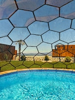 Piscina com cobertura de vidro hexagonal, cercada por gramado e cerca de madeira.