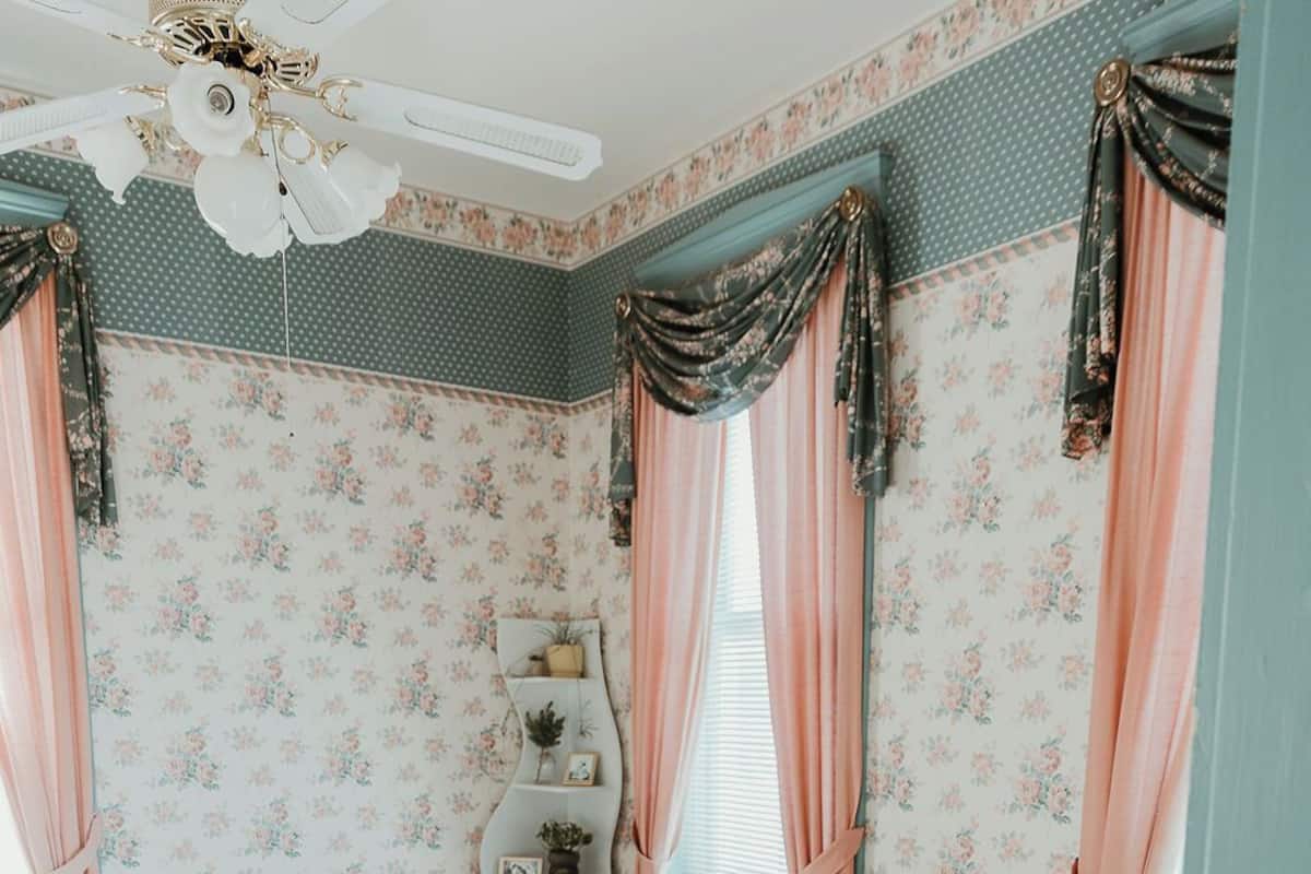 Uma sala de estar com papel de parede floral, um sofá bege, um banquinho, um ventilador de teto e uma janela com cortinas.