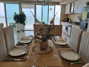 Uma zona de refeições com vista para o mar, uma cozinha com eletrodomésticos modernos e uma mesa de jantar posta com pratos e copos.