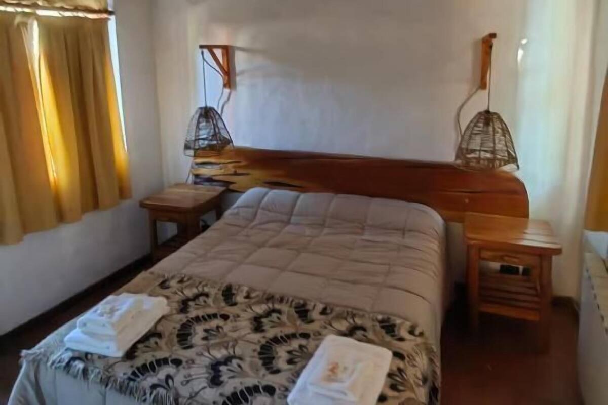 Quarto com cama, cabeceira de madeira, mesas de cabeceira, luminária e janela com cortinas.
