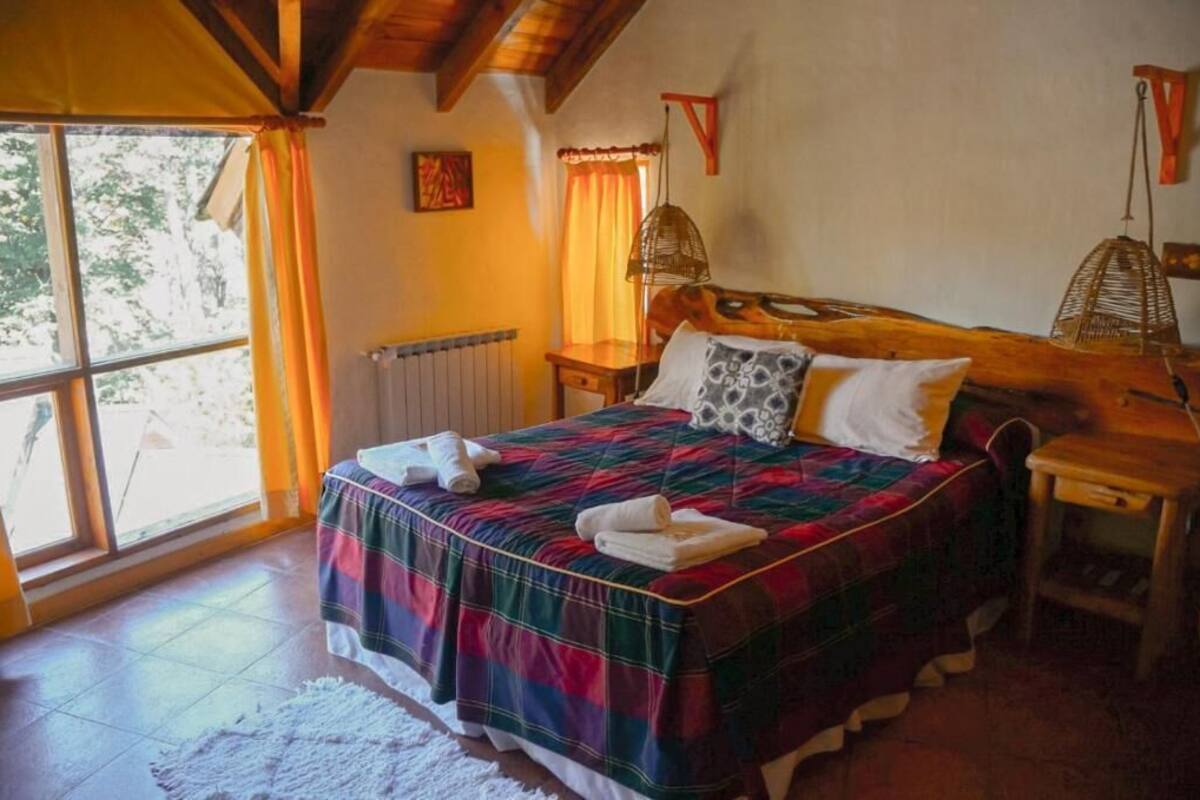 Quarto com cama, cabeceira de madeira, janela com cortinas, radiador e luminária pendente.