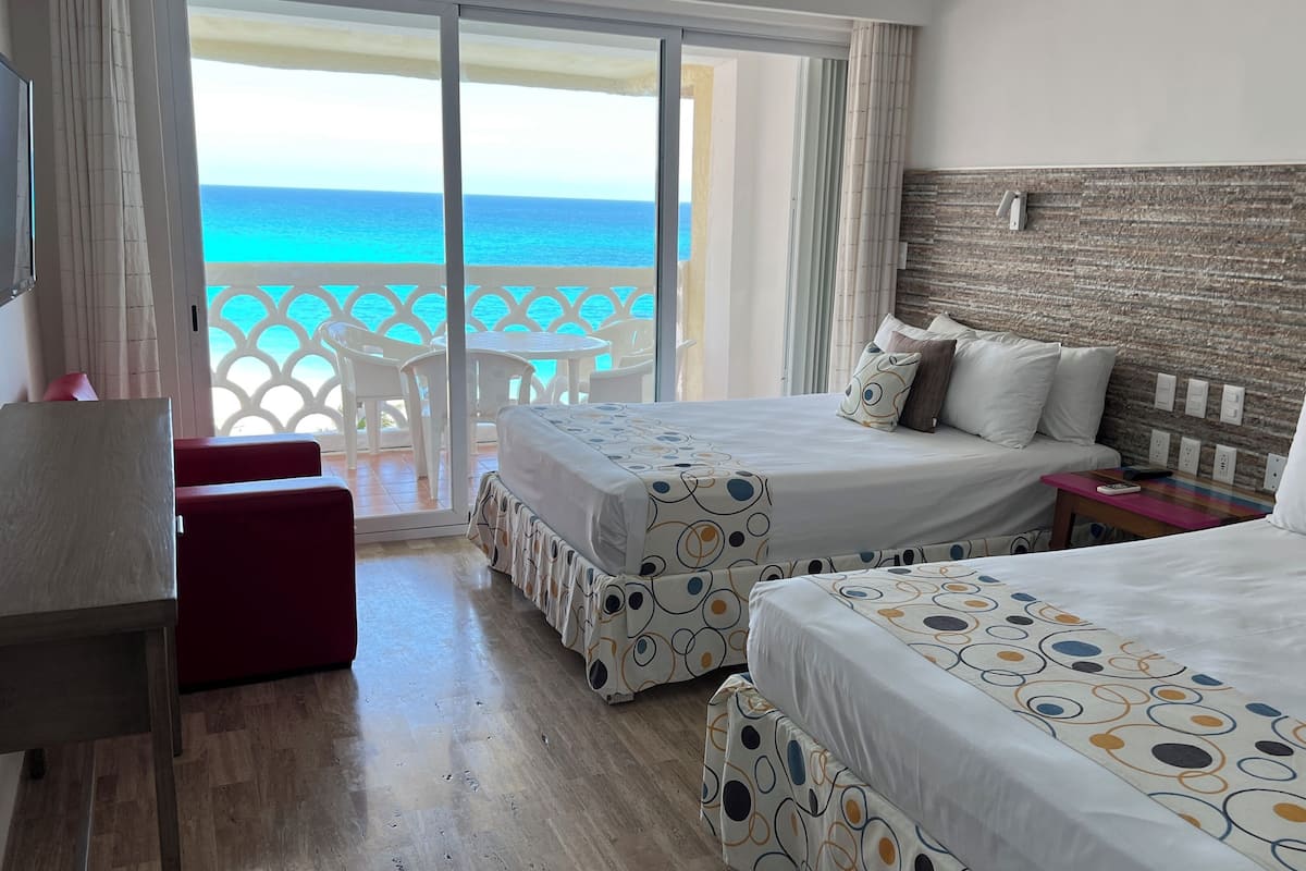 Quarto de hotel com duas camas, uma escrivaninha, uma cadeira e varanda com vista para o mar.