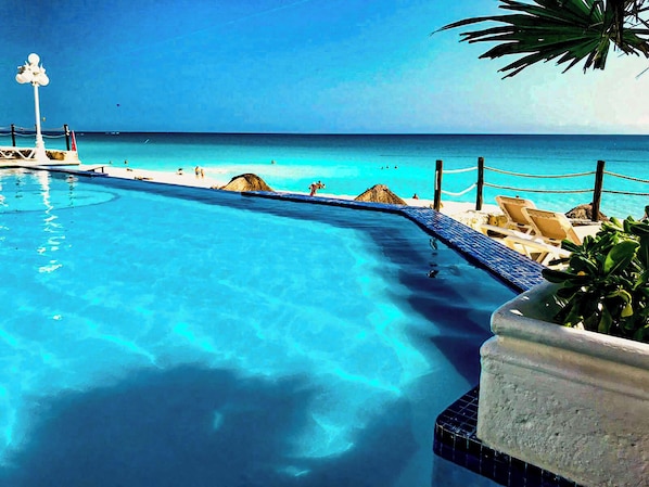 Uma piscina azul cristalina com vista para o oceano, cadeiras de descanso e uma estrutura decorativa branca.