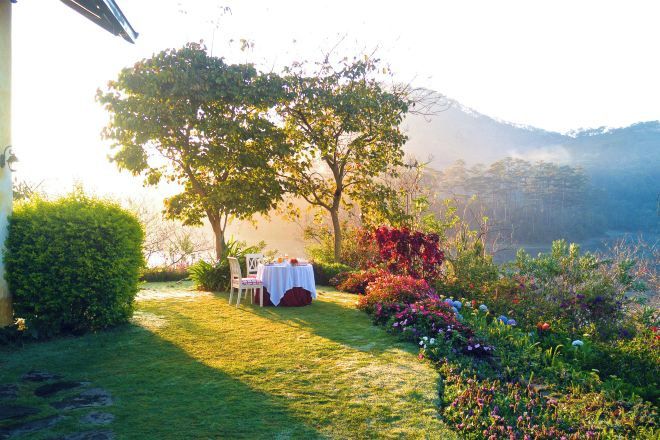 Uma área de refeições ao ar livre com mesa e cadeiras, rodeada por vegetação exuberante e plantas floridas.