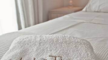Uma cama bem arrumada com uma toalha branca, uma luminária na mesa de cabeceira e uma janela com cortinas.