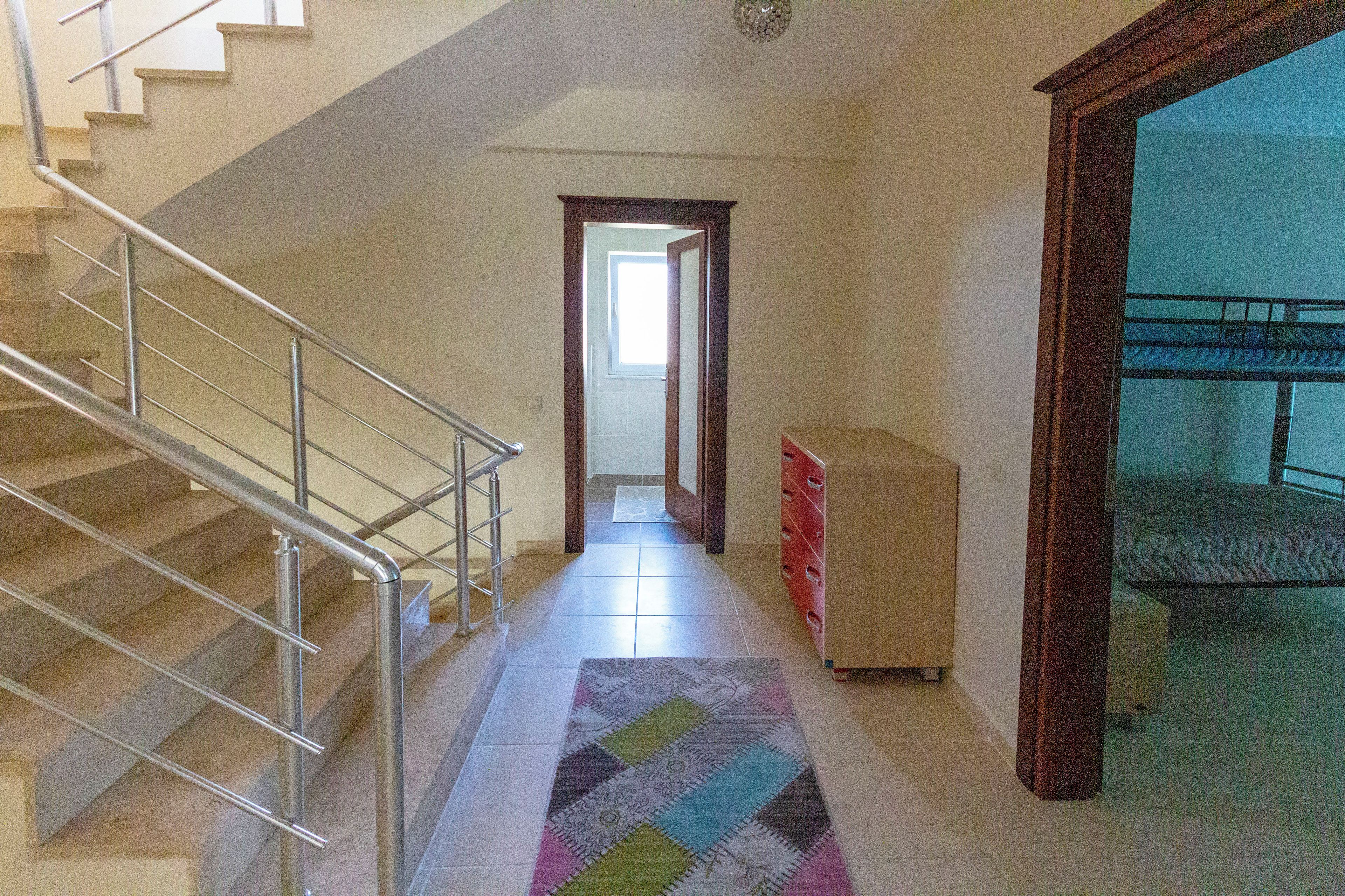 Uma escada com guarda-corrimão de metal leva a um quarto com beliche. Um tapete colorido está no chão, e há um armário de madeira no quarto.
