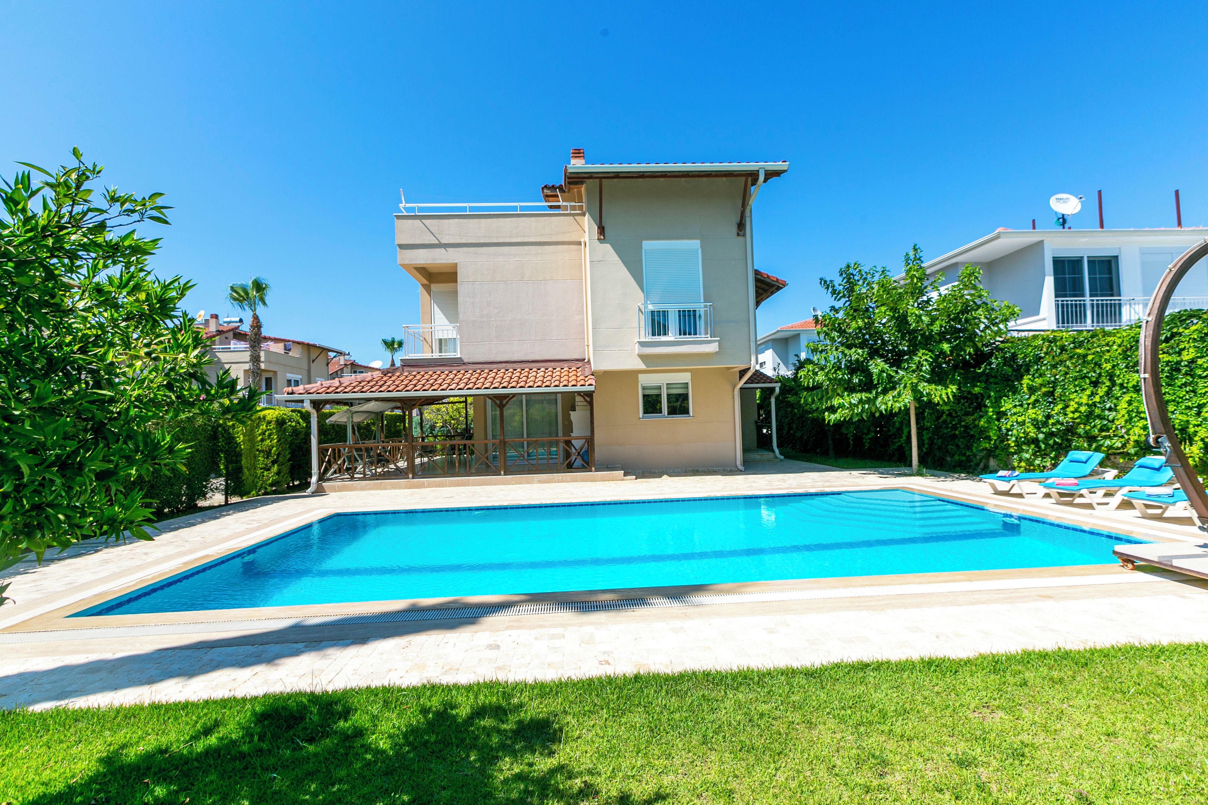 Uma casa moderna de dois andares com piscina, rodeada por vegetação e outras casas.