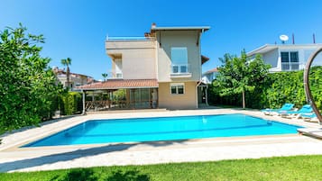 Uma casa moderna de dois andares com piscina, rodeada por vegetação e outras casas.