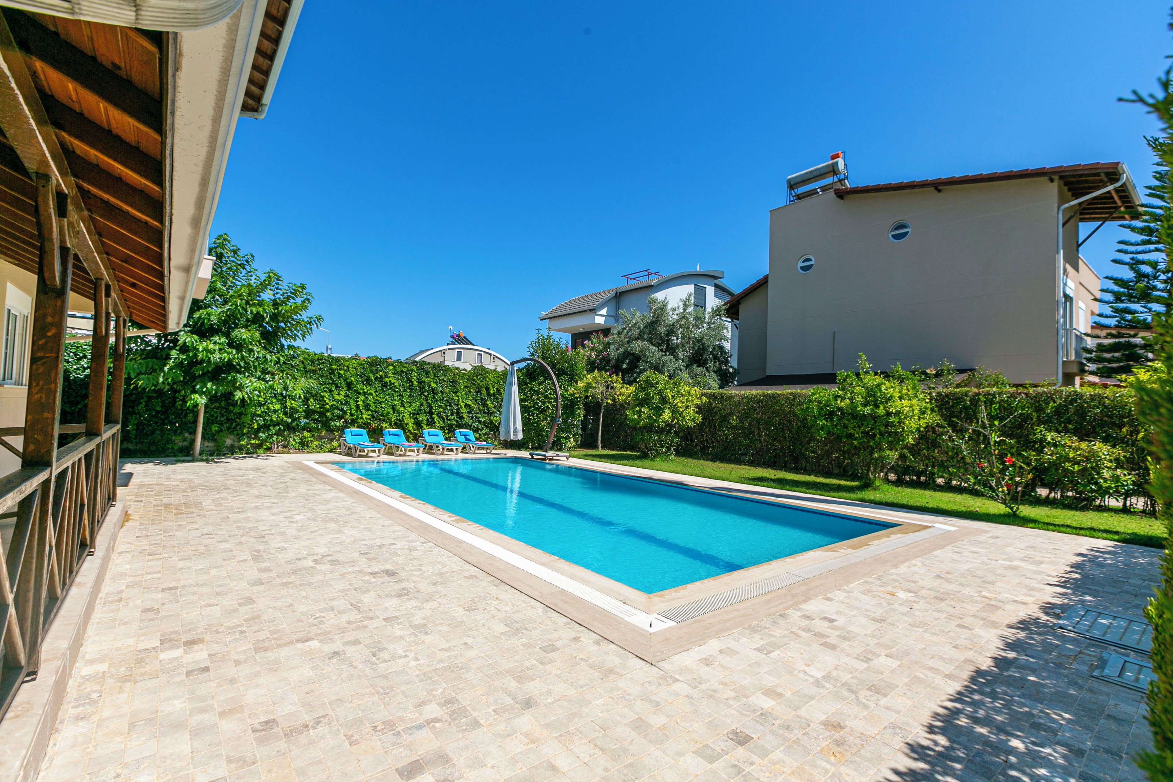 Piscina com um deck revestido em azulejos, rodeada por vegetação e edifícios modernos.