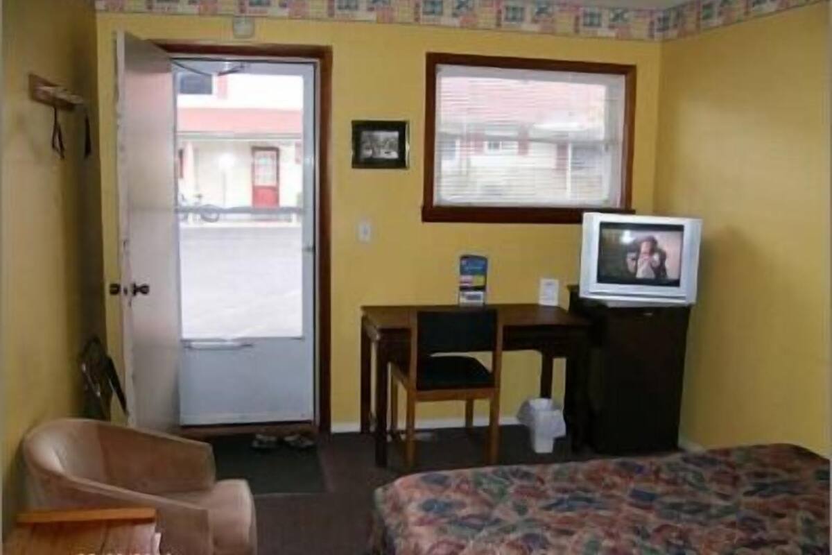 Quarto com cama, escrivaninha com televisão, cadeira e porta que dá para o exterior.