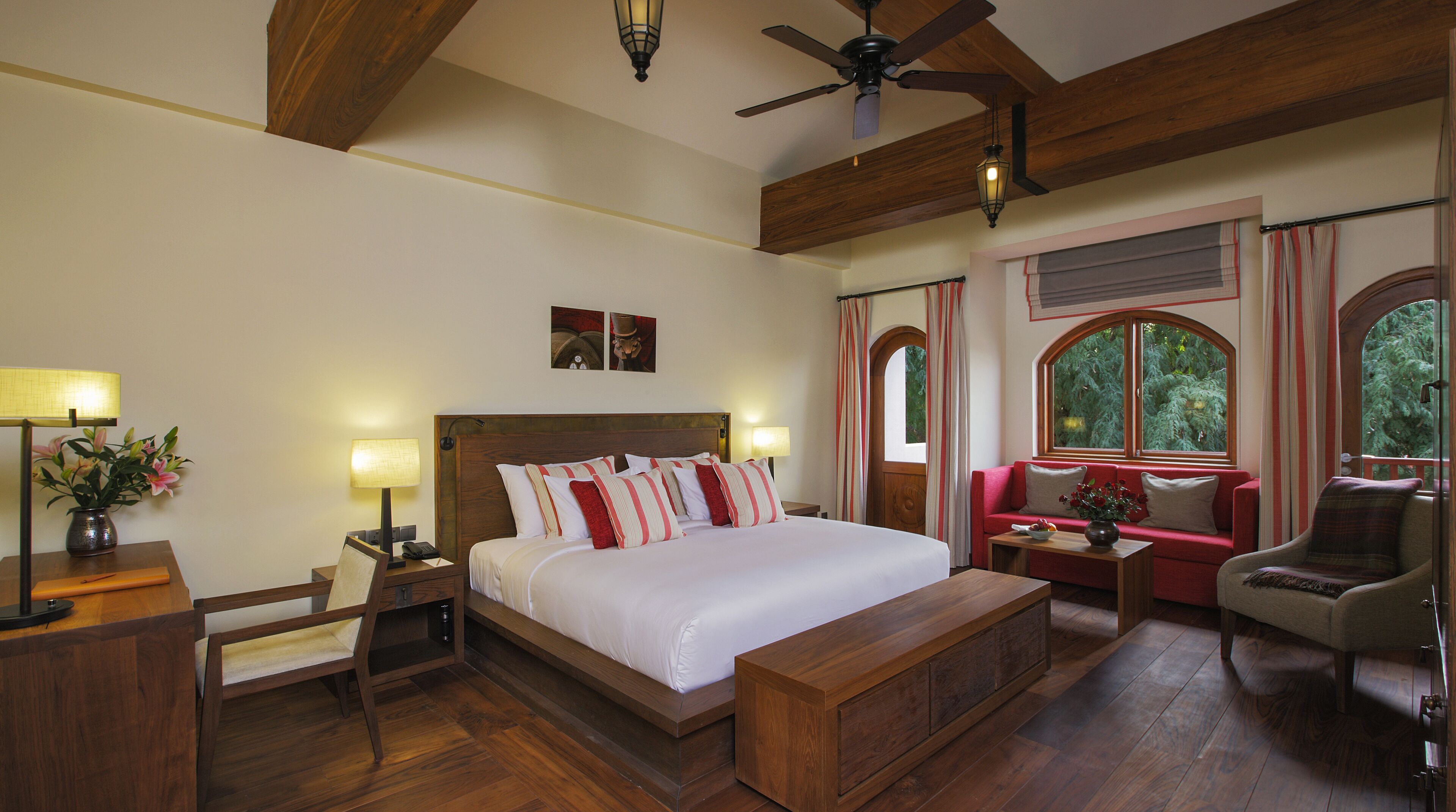 Quarto de hotel com uma cama grande, cabeceira de madeira, escrivaninha com luminária, cadeira, sofá e janela com cortinas.