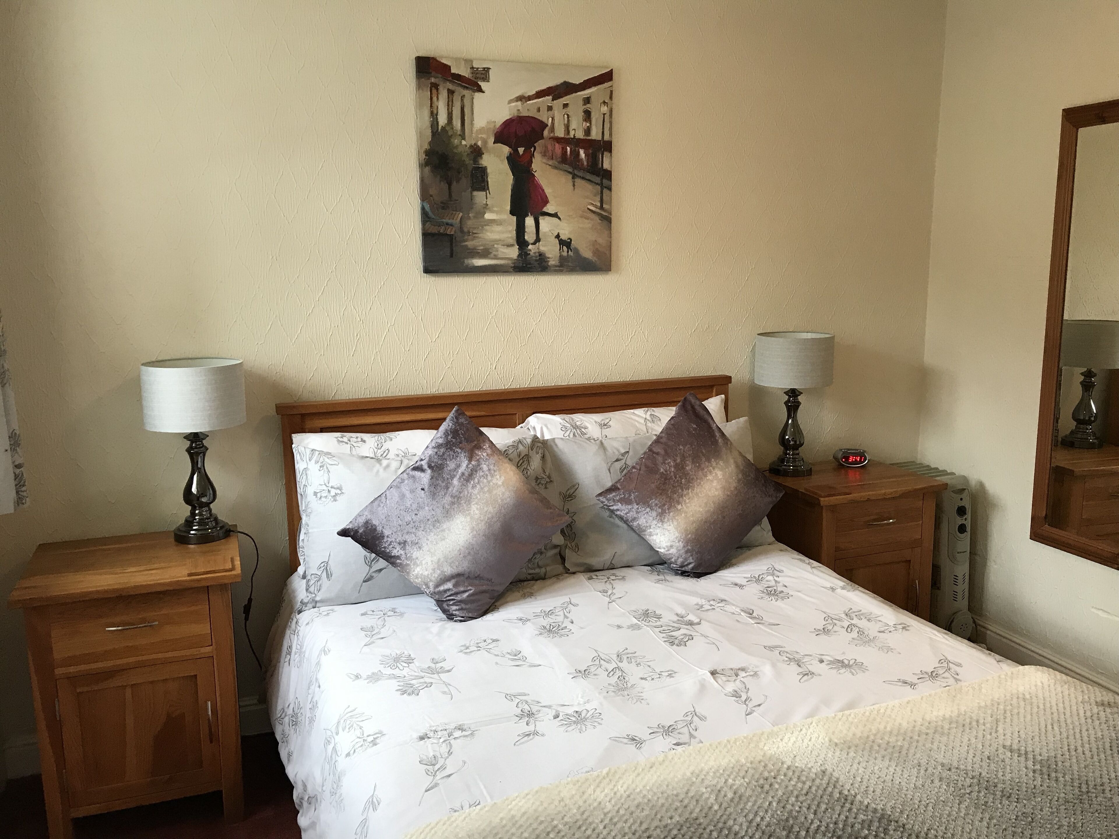 Um quarto com cama, duas mesas de cabeceira, um criado-mudo, abajur, relógio, espelho e um quadro na parede.
