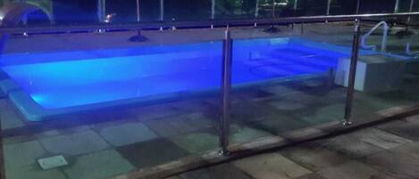 Piscina externa com barreira de vidro e piso azulejado, cercada por guarda-sóis e uma palmeira.