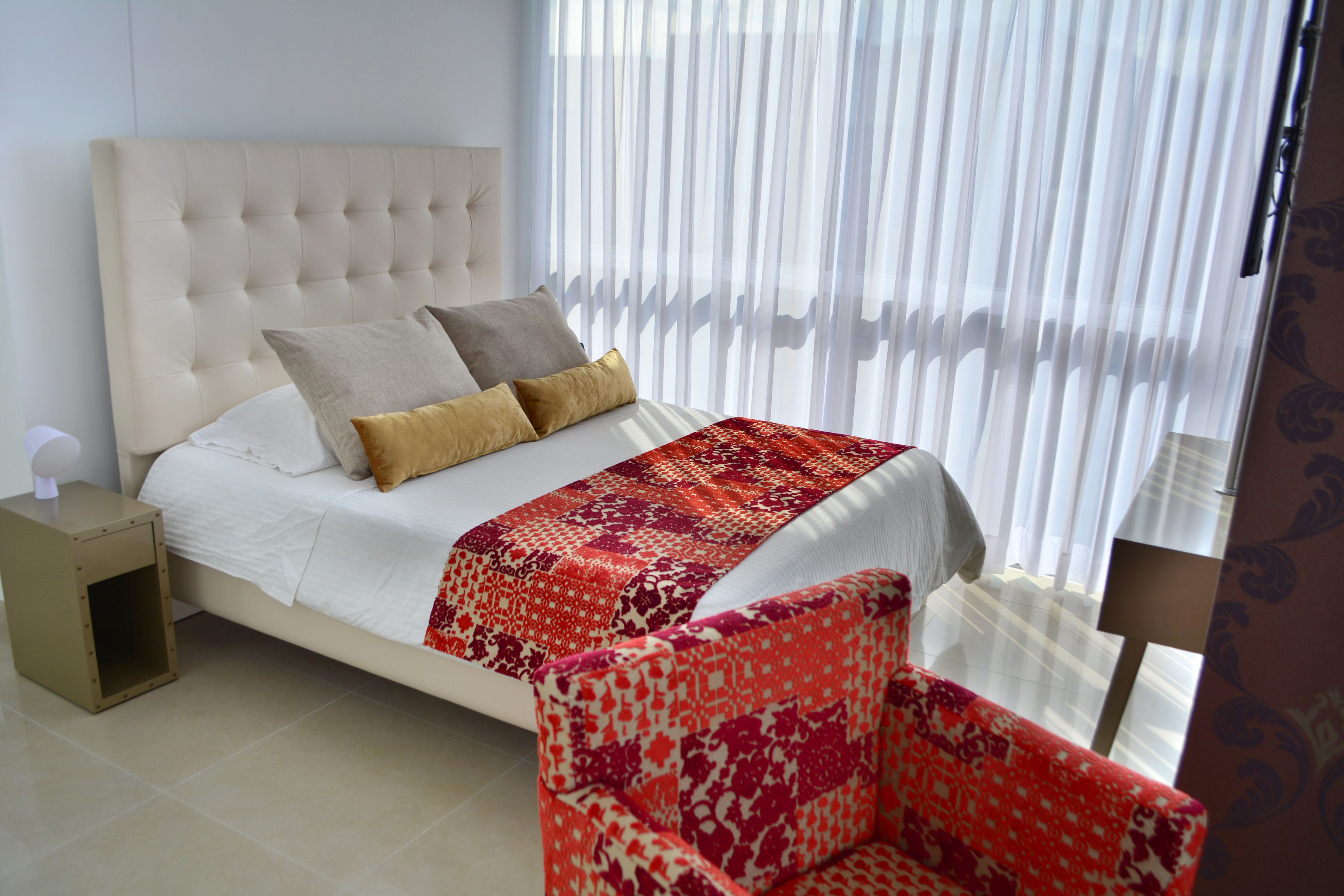 Quarto com uma cama grande, uma mesinha de cabeceira, uma cadeira e uma janela com cortinas leves.