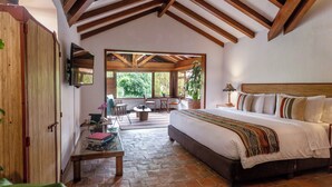 Quarto com cama grande, vigas de madeira, televisão, escrivaninha com cadeira e vista para o exterior.