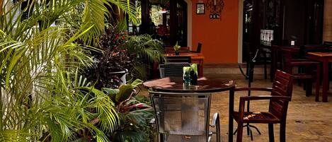 Um pátio ao ar livre com mesas e cadeiras, rodeado por plantas em vasos e um mural na parede.