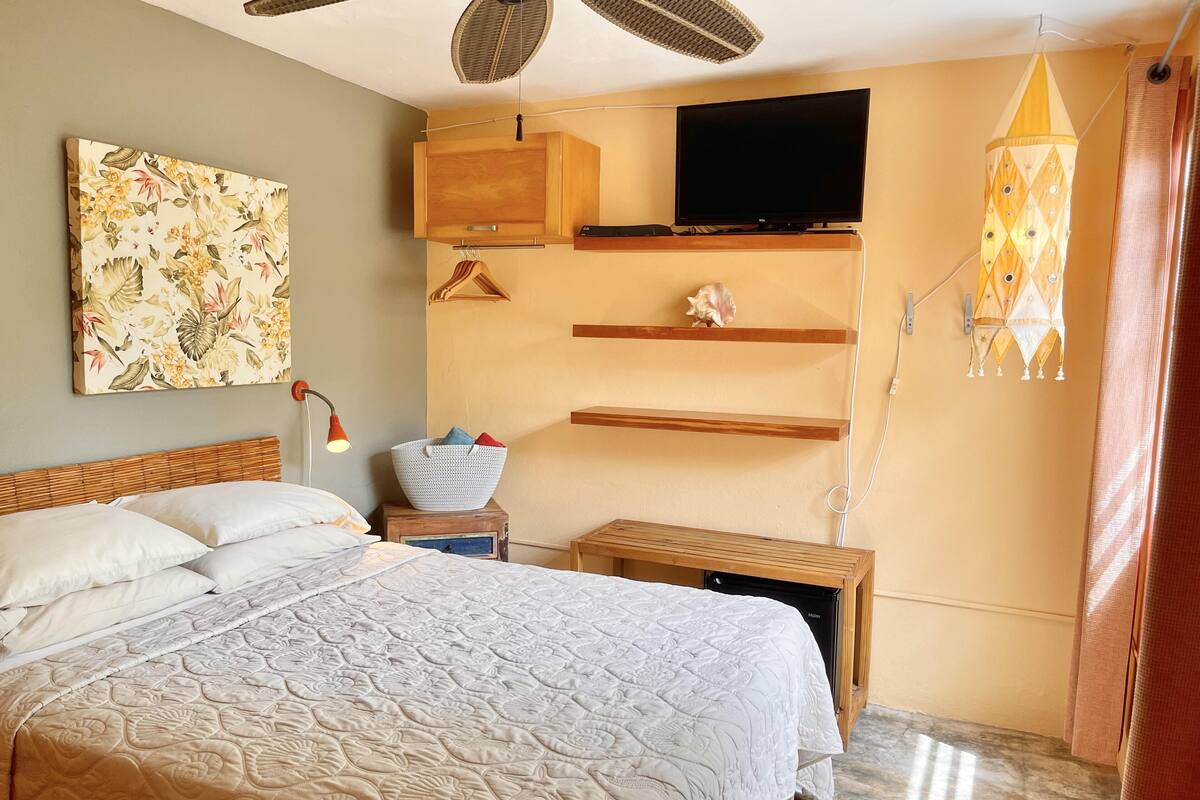 Quarto com cama, televisão, prateleira na parede, luminária pendente e um objeto decorativo na parede.