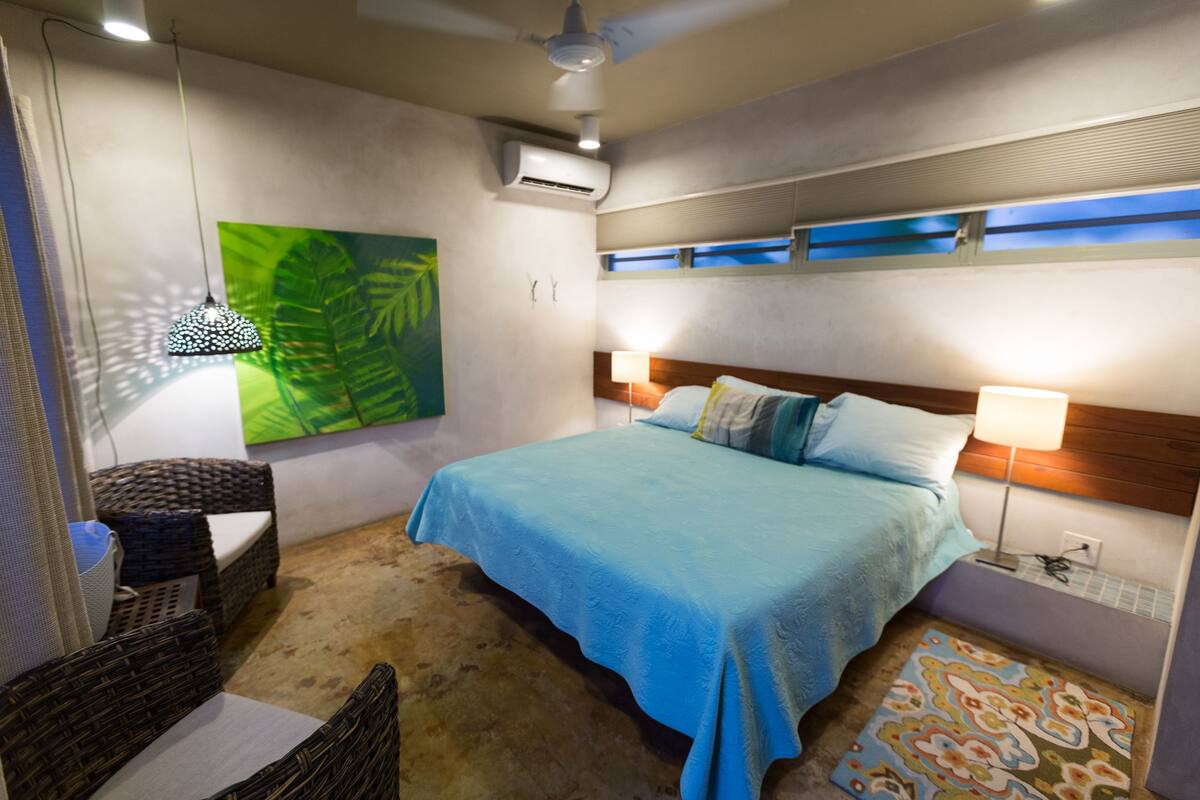 Um quarto com cama, duas luminárias de cabeceira, ar-condicionado fixado na parede, ventilador de teto, pintura de uma folha verde grande e móveis de vime.