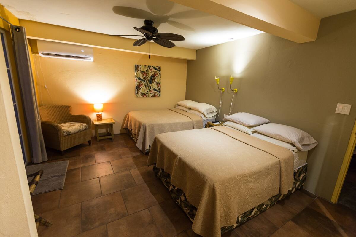 Quarto de hotel com duas camas, um ventilador de teto, aparelho de ar condicionado, um quadro na parede e mesa de cabeceira com abajur.
