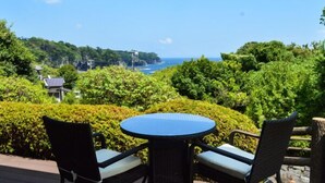 Um pátio com uma mesa redonda e duas cadeiras, com vista para uma paisagem costeira com vegetação e o mar.