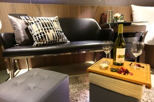 Uma sala de estar moderna com um sofá de couro preto, um banquinho branco e uma mesa de centro de madeira com uma garrafa de vinho, copos e uma vela.