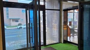 Entrada de hotel com portas de vidro, gramado artificial verde e vista para a rua e edifícios.