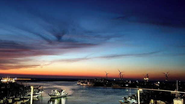 Uma marina ao pôr do sol com barcos atracados, uma paisagem urbana ao fundo e turbinas eólicas.