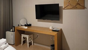 Quarto de hotel com escrivaninha de madeira, televisão, pequeno frigorífico, mesa de cabeceira com candeeiro e cama.