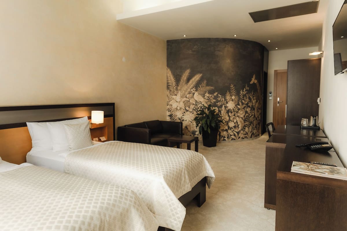 Quarto de hotel com duas camas, uma escrivaninha, uma cadeira, um vaso de plantas e um grande mural na parede.