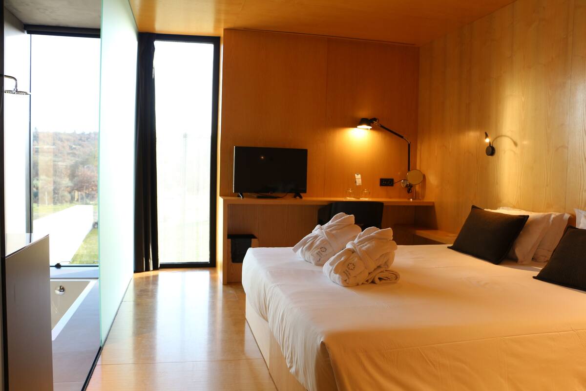 Quarto de hotel com cama, televisão, escrivaninha e vista para o exterior.
