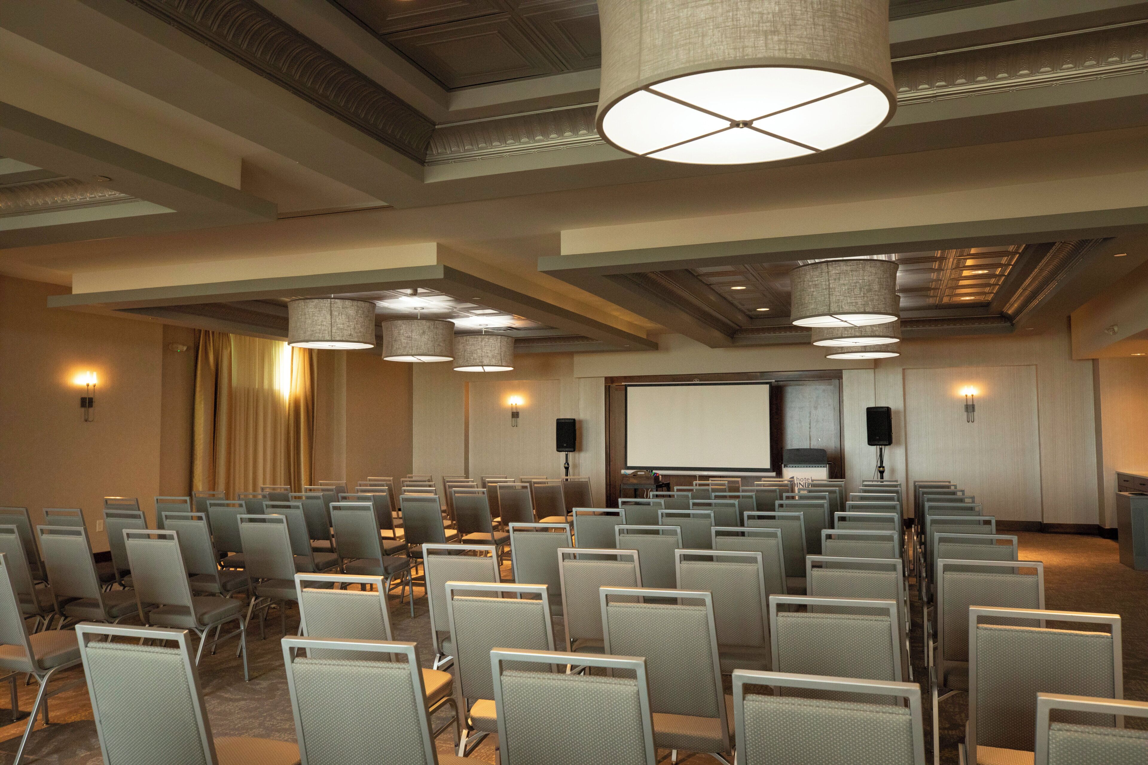 Uma sala de conferências com fileiras de cadeiras, uma tela de projetor e luminárias no teto.