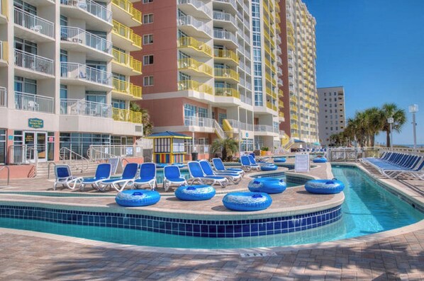 Área de piscina de hotel com espreguiçadeiras azuis e boias infláveis, cercada por um edifício com várias andares e varandas.