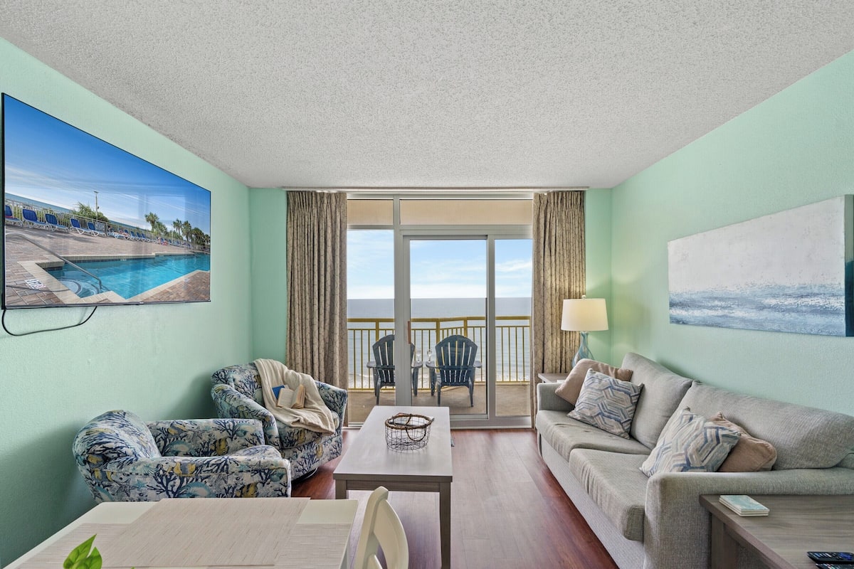 Quarto de hotel com vista para a piscina, varanda com cadeiras e um sofá.