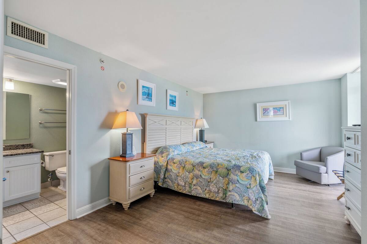 Um quarto com cama, mesas de cabeceira, uma cadeira e um armário.