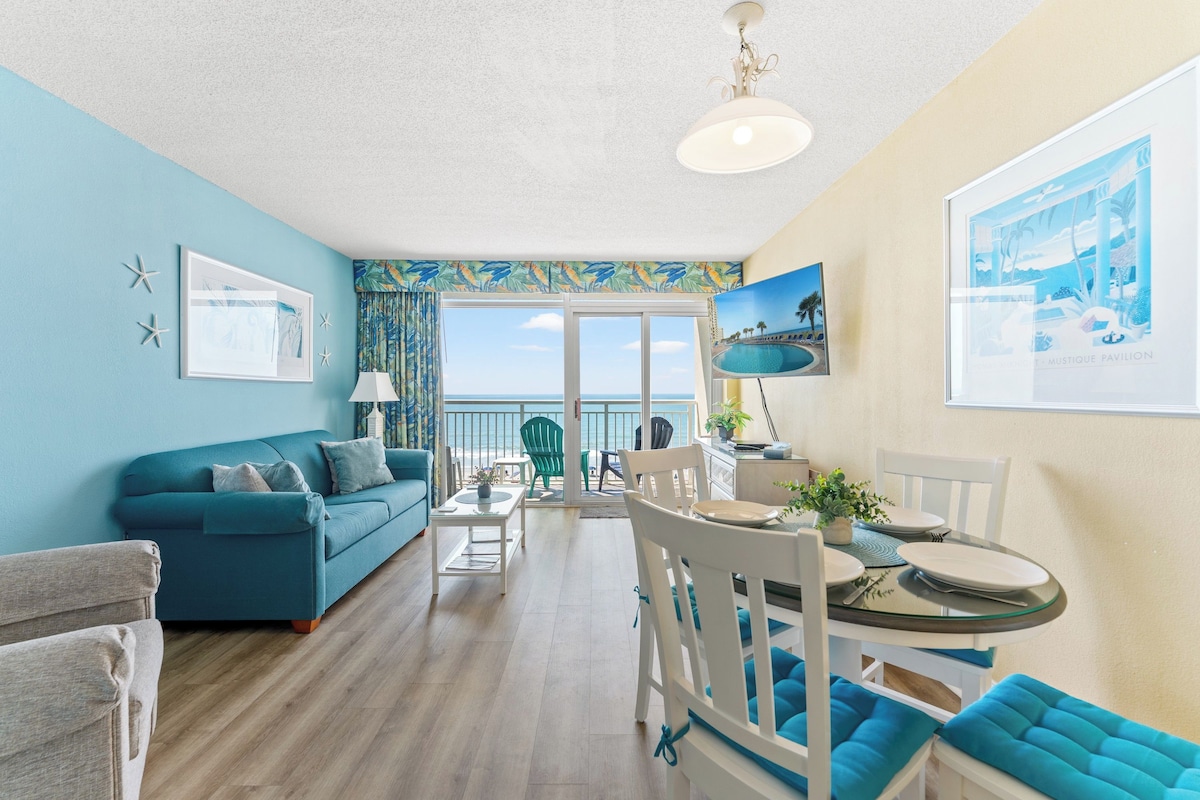 Quarto com sofá azul, mesa de jantar com cadeiras, vista para o oceano e decoração com tema tropical.