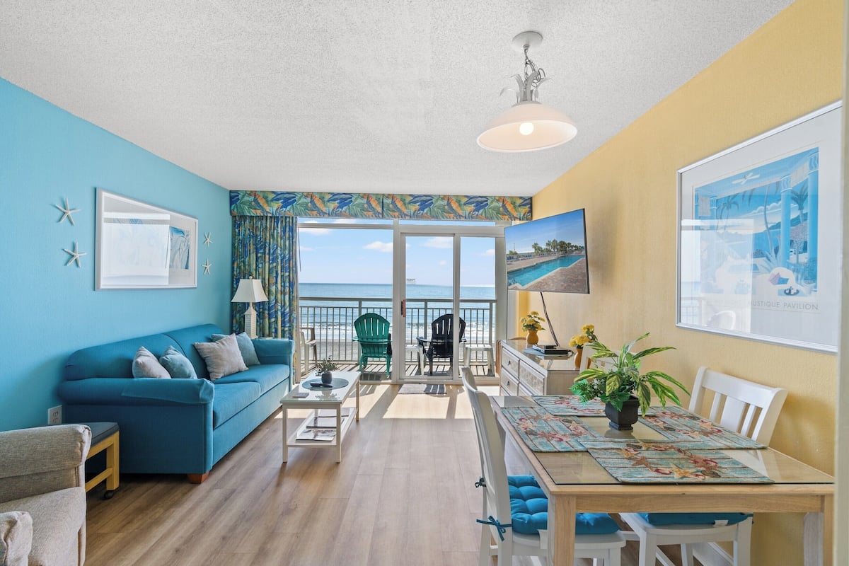 Uma sala de estar com sofá azul, uma mesa de jantar com cadeiras, uma varanda com vista para o mar e um quadro na parede.