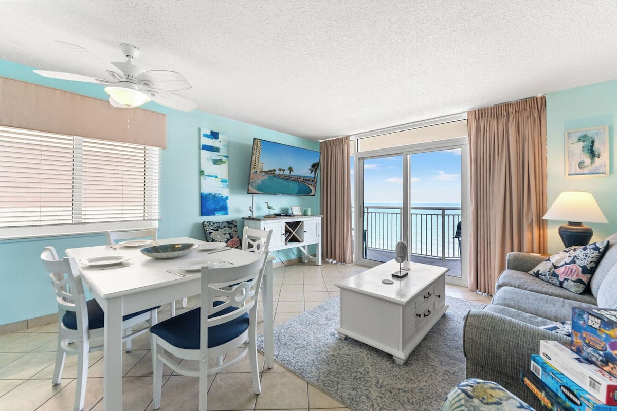 Quarto com uma mesa de jantar, cadeiras, um armário branco, um sofá, uma televisão e vista para o oceano.